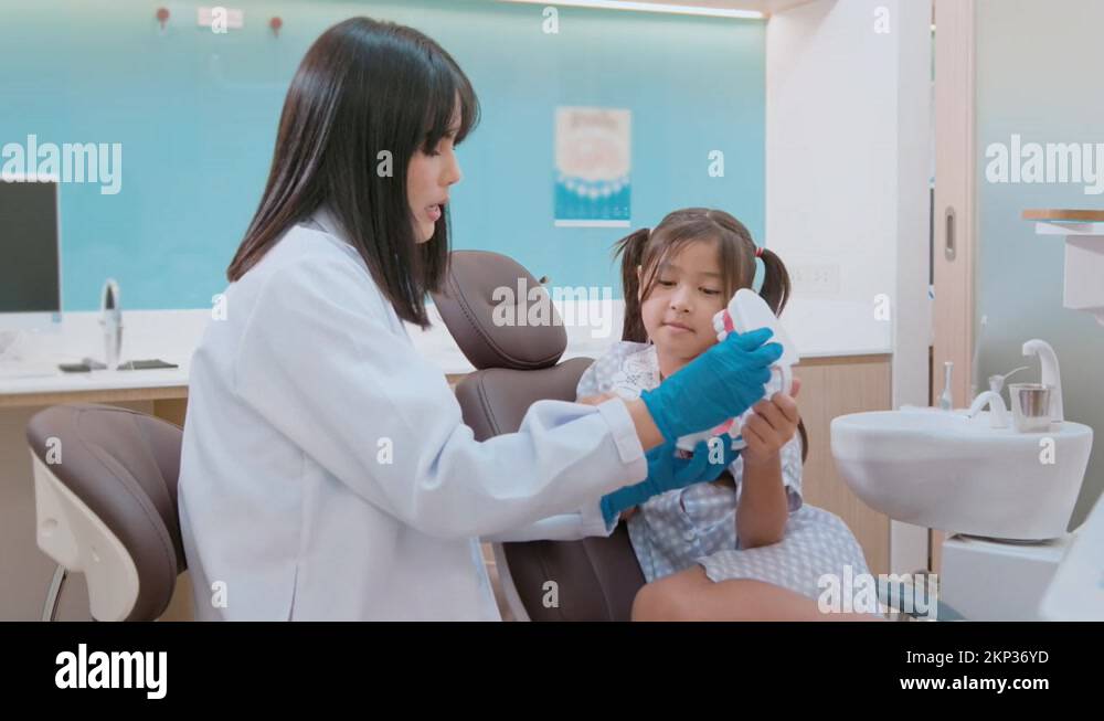 Female dentist demonstrating how to brush teeth to a little girl in ...
