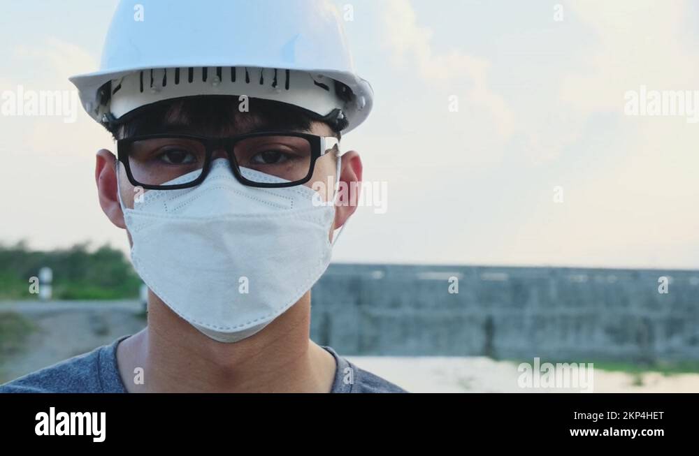 Young Asian engineer wearing a helmet and mask looks and smiles at the ...