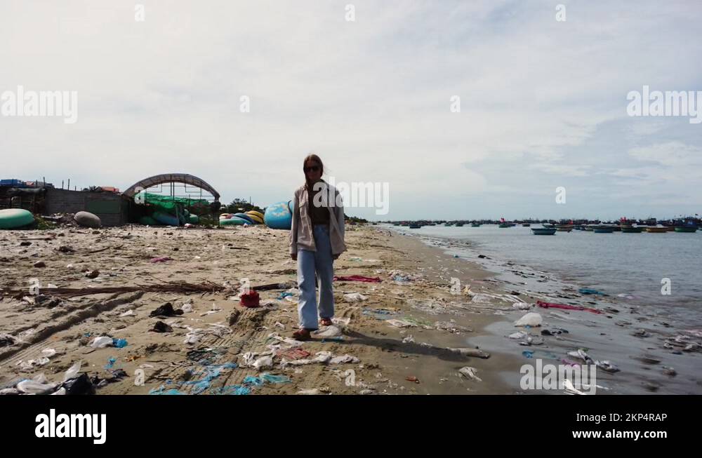 Young woman walking alone on polluted beach with plastic waste wearing ...