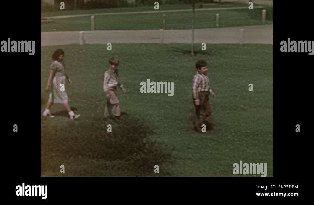 1950s: children walk in single file across grass in front of school ...