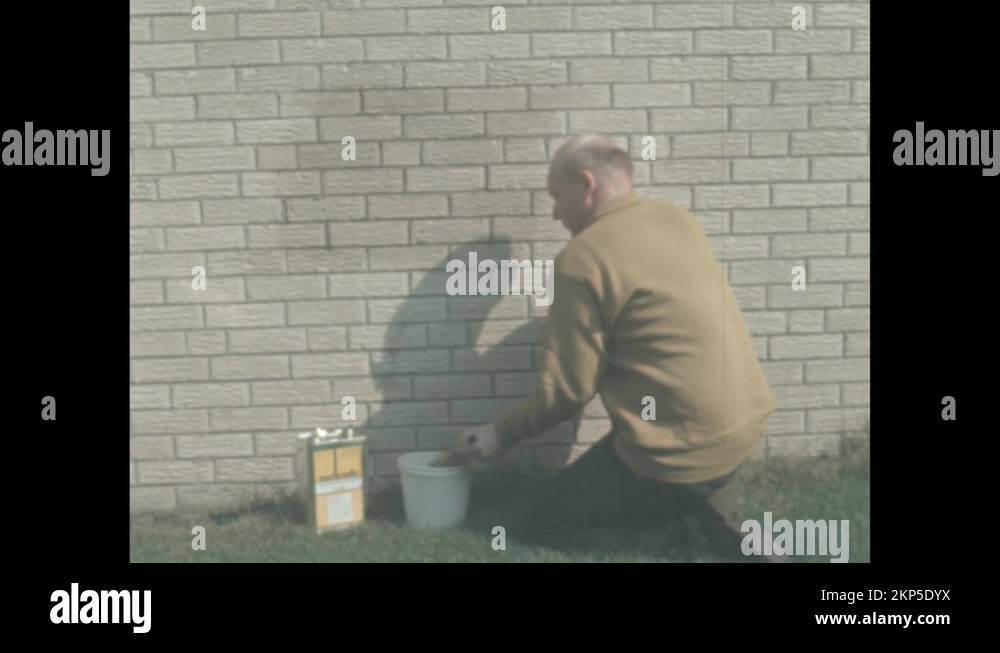 1960s: Man kneeling, painting brick wall. Man running paint brush over ...