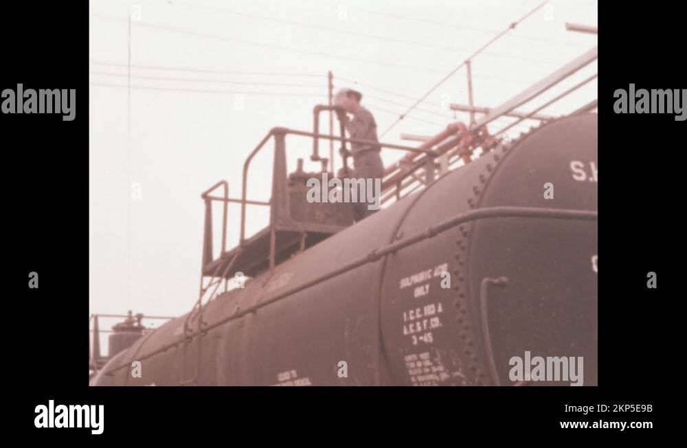 1960s: Man walks on top of tanker car, attaches pipe to top of car ...