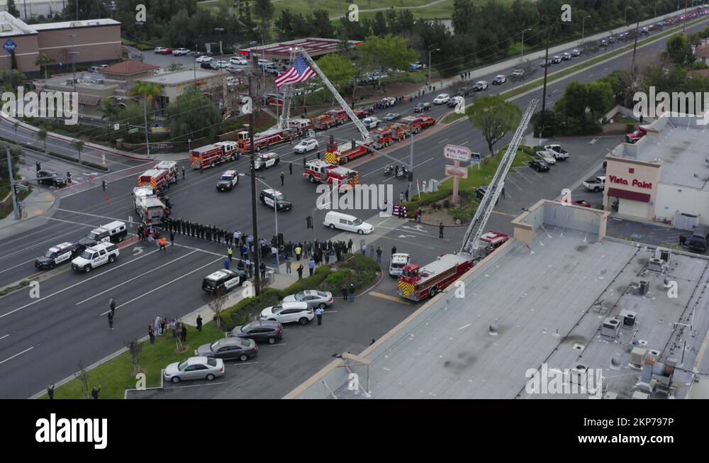 A funeral procession lead by a white hearse drives past a community ...