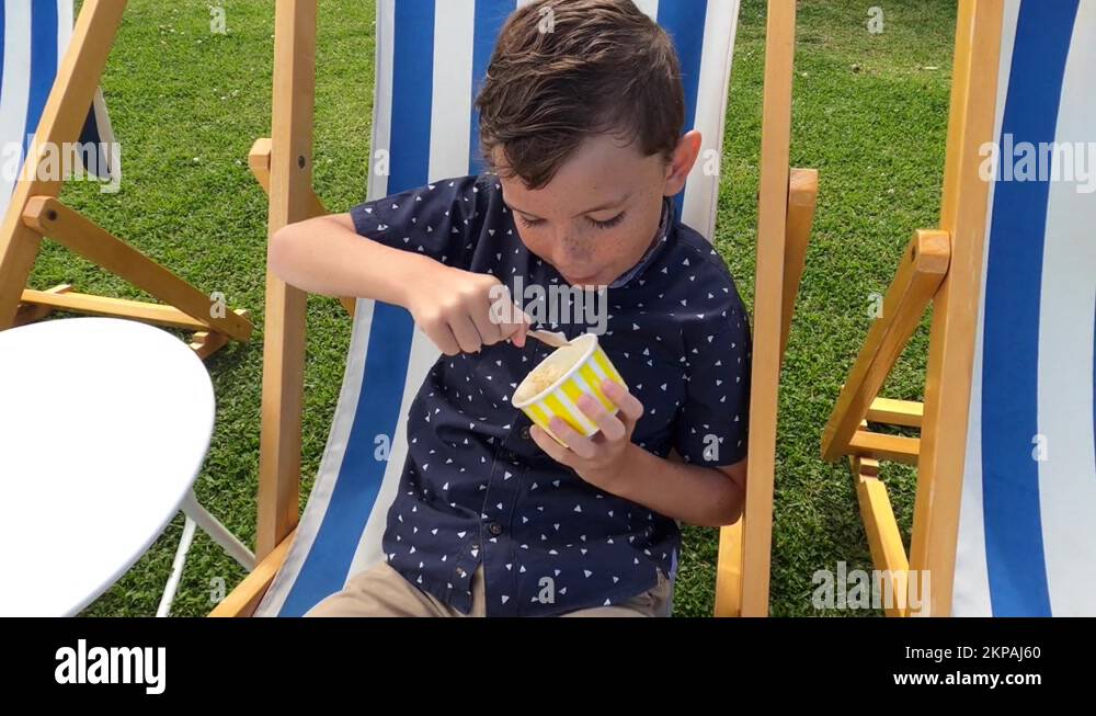 A smartly dressed boy eats ice cream from a tub, sitting on a deck ...