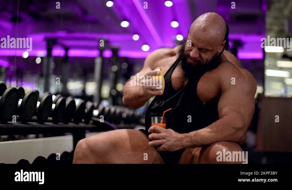 A brutal male bodybuilder sits in the gym and squeezes fruit into a ...