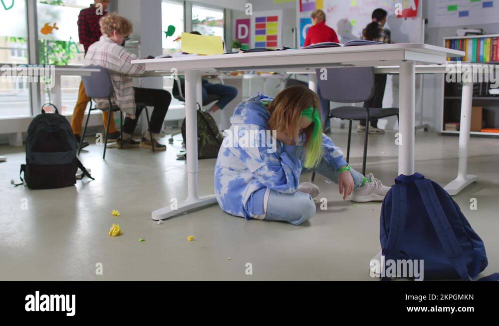 Bullying at school. Stressed teen girl sit under desk in classroom and ...