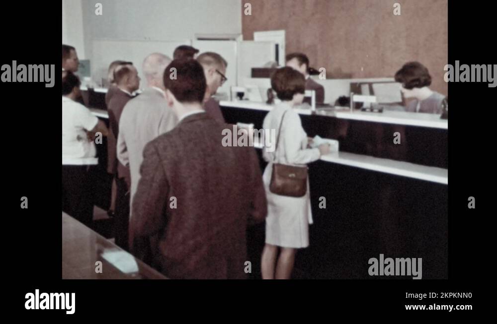 1960s: Customers and tellers in bank lobby. Teller helps customer. Man ...