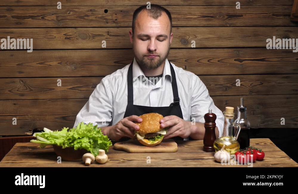 bearded young male chef eats homemade burger. Junk food, modern life ...