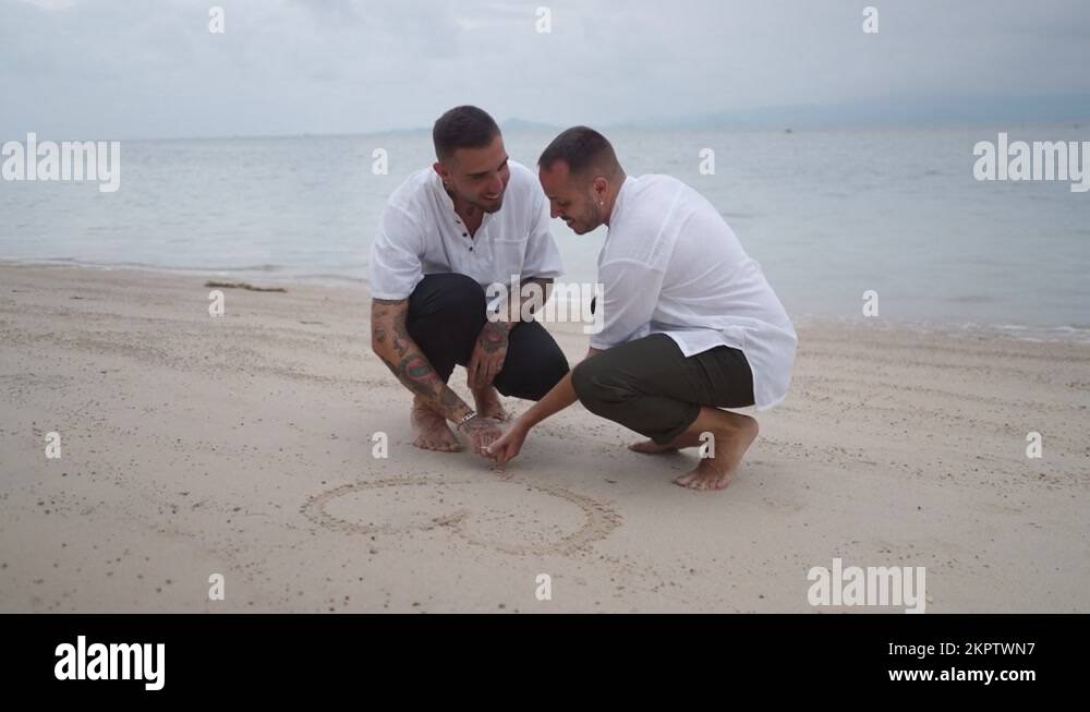 Engaged Gay couple honeymooners draw a heart shape on the sand and kiss ...