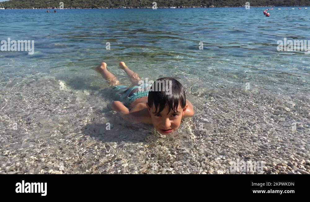 Boy rests on his belly on the pebbles in the crystal clear sea waters 2 ...
