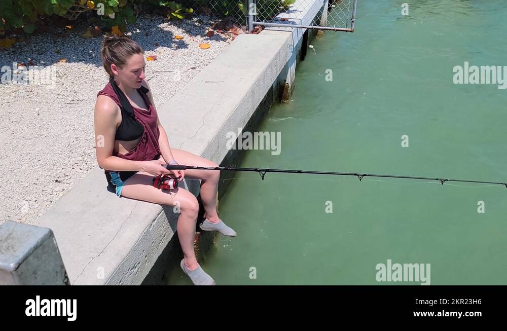 Woman Fishing while Sitting on Edge of Pier on Shore while she Reels ...