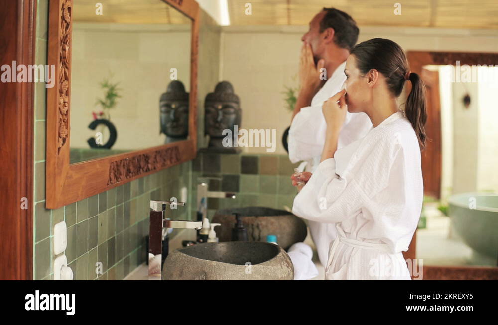 Woman cleaning face with cotton pad, husband applying creme in bathroom ...