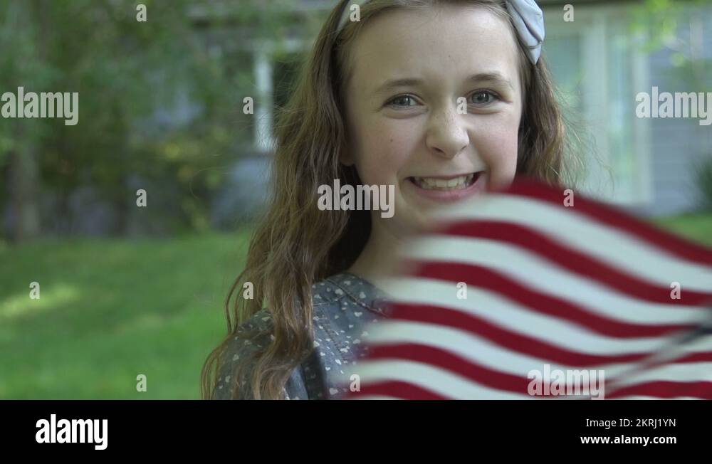 A tween girl smiles at camera in front of her house holding a US flag ...