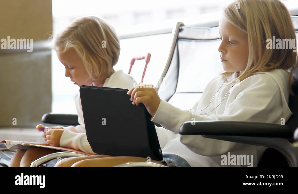Two young girls sit in chairs waiting while playing on a tablet and ...