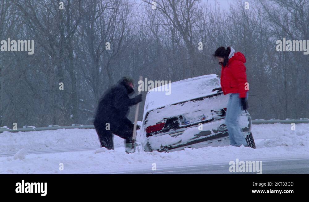 Man digging out a car stuck in snow beside a highway while woman looks ...