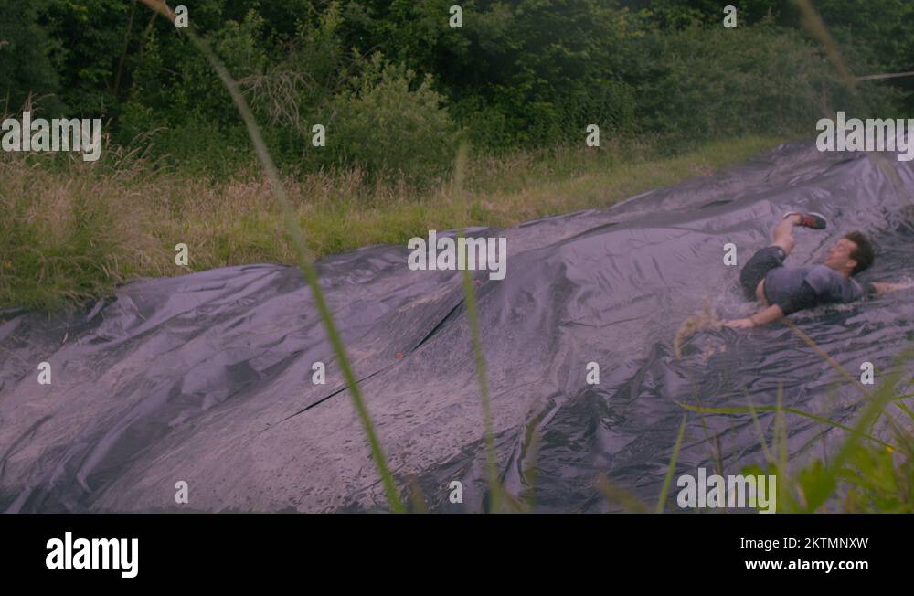 4K Mud race runner on assault course slides down hill into cold muddy ...
