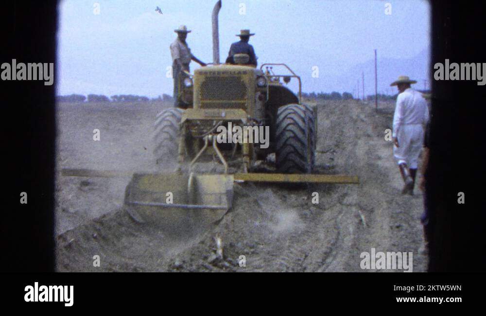 1965: a man driving a large backhoe to make a ditch as men walk along ...