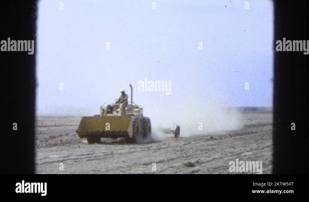 1965: workman driving a big machine on a dusty road in a deserted area ...