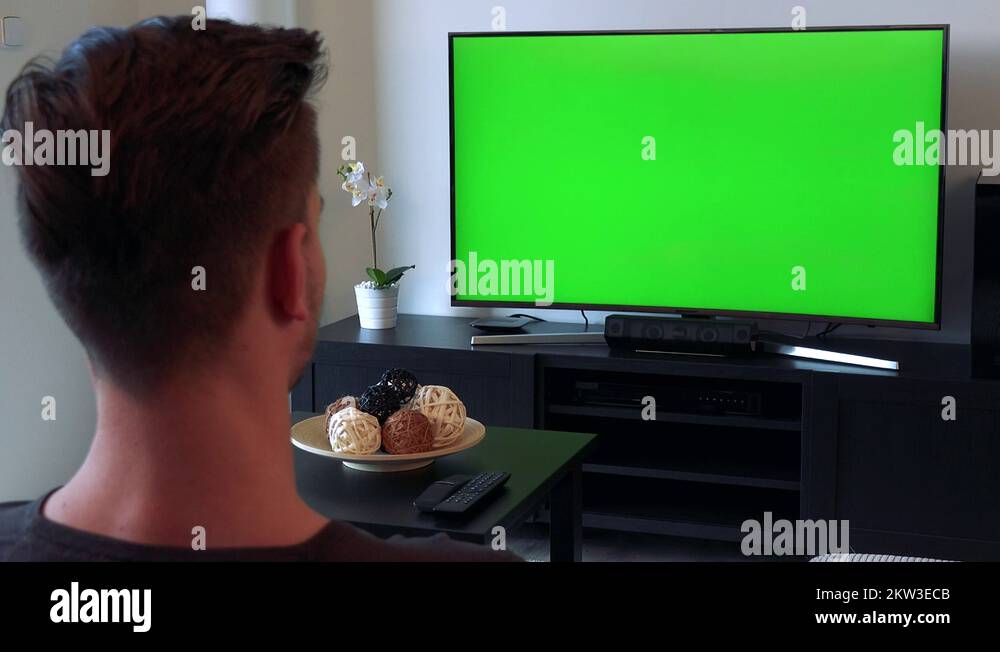 A man, the back of the head to the camera, watches a TV with a green ...