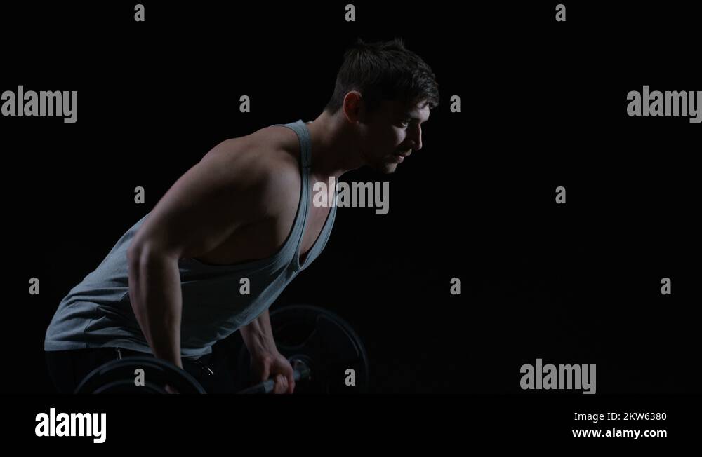 4K Fit young man with muscular physique lifting weights against black ...