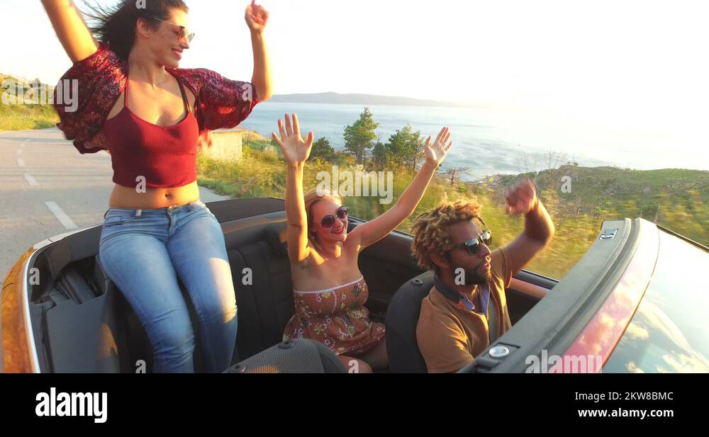 Beautiful young brunette sitting on hood of convertible and dancing to ...