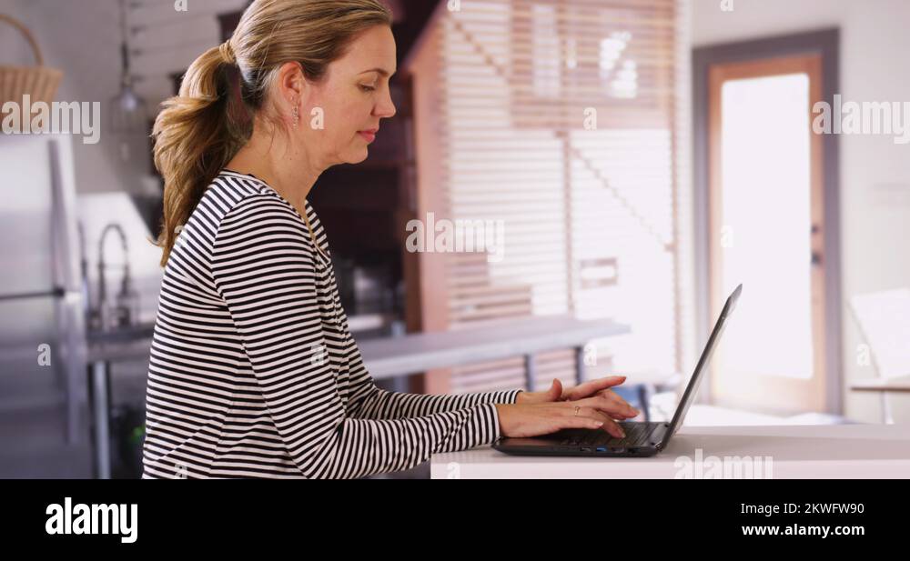 Profile of Caucasian woman seated a counter in modern kitchen typing on ...