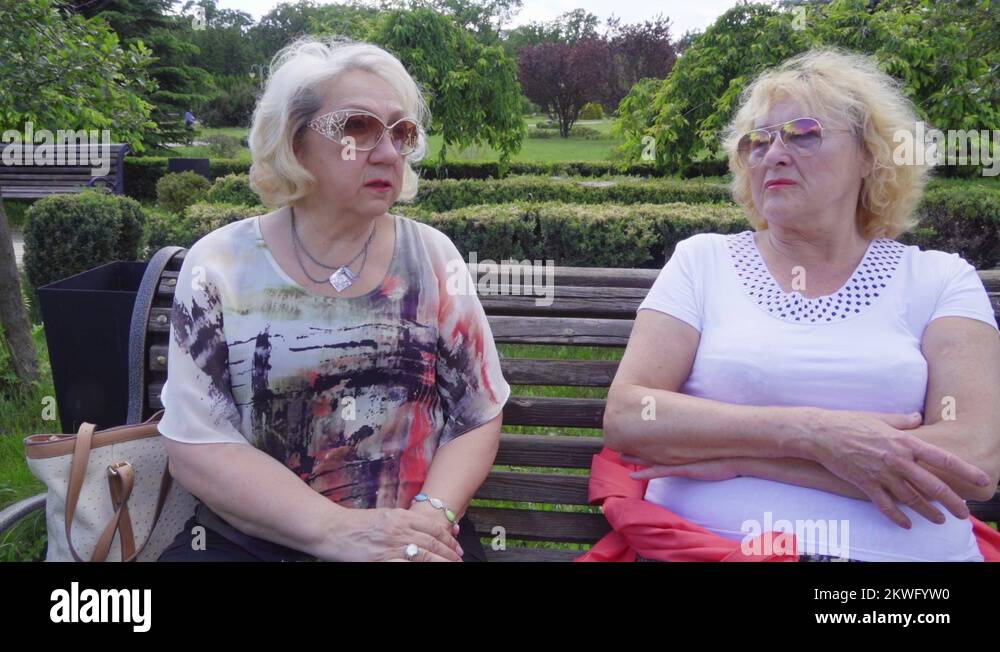 Two sad senior women sit on a park bench and shake their heads sadly ...