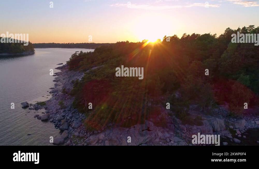 Drone flying forward towards sun at rocky shore over island with forest ...