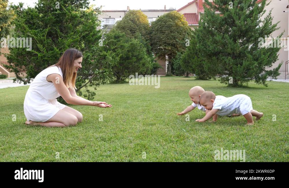 Two babies twins boys crawling to their young mother over the lawn ...