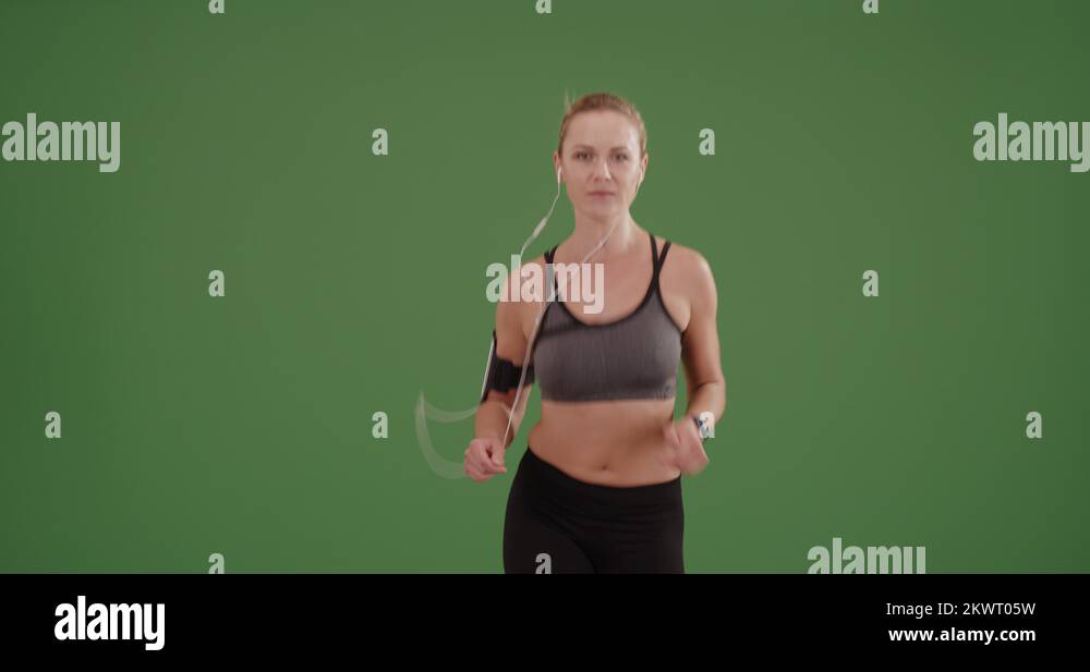 Caucasain woman with earbuds in running toward the camera on green ...