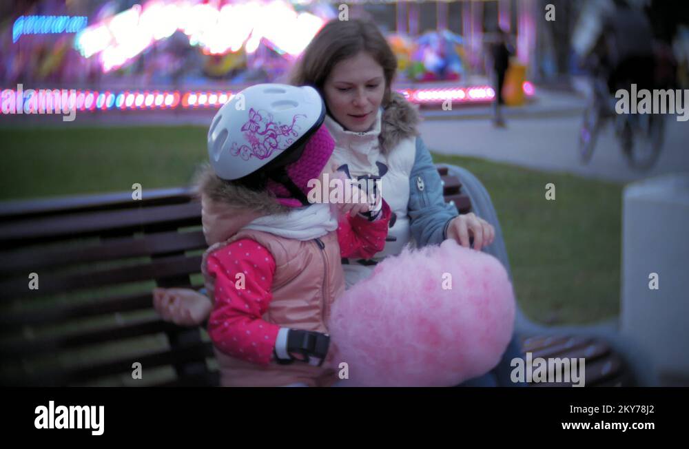 Beautiful little girl sitting on a bench in an amusement park, eating ...