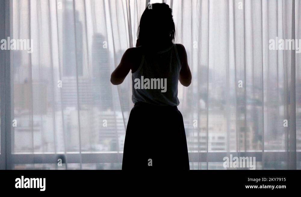 Silhouette of woman unveil curtains and admire city form the window ...