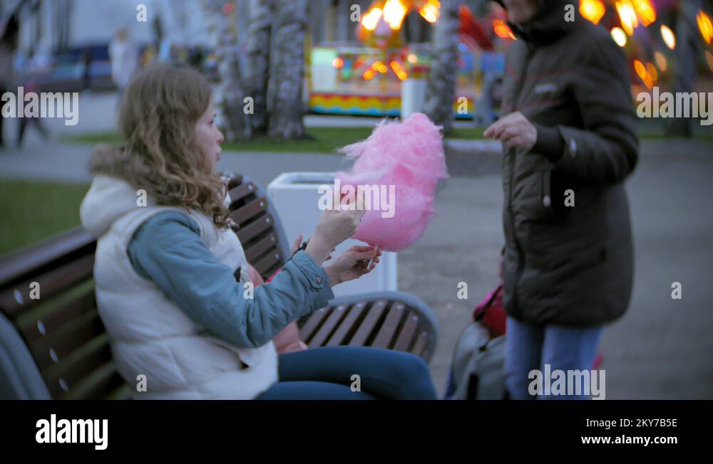 Beautiful little girl sitting on a bench in an amusement park, eating ...