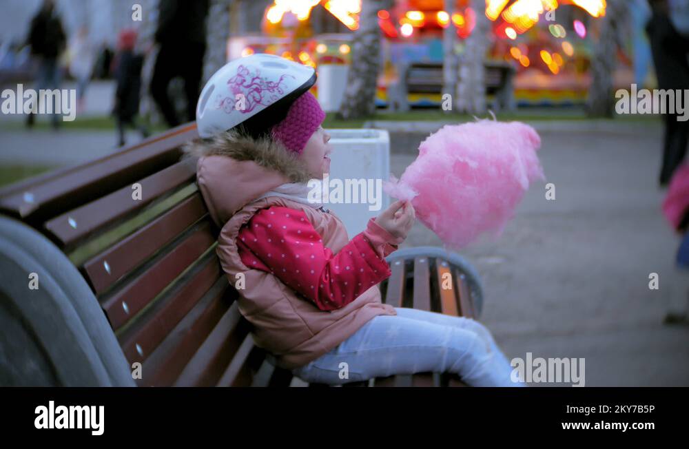 Beautiful little girl sitting on a bench in an amusement park, eating ...
