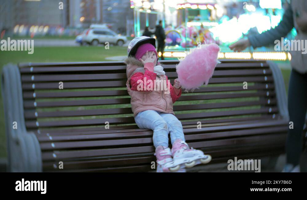 Beautiful little girl sitting on a bench in an amusement park, eating ...