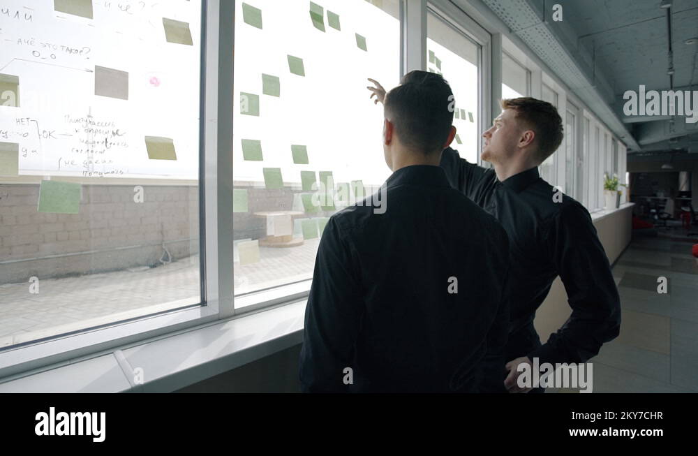 Young men are talking, pointing on notes on window in modern office ...