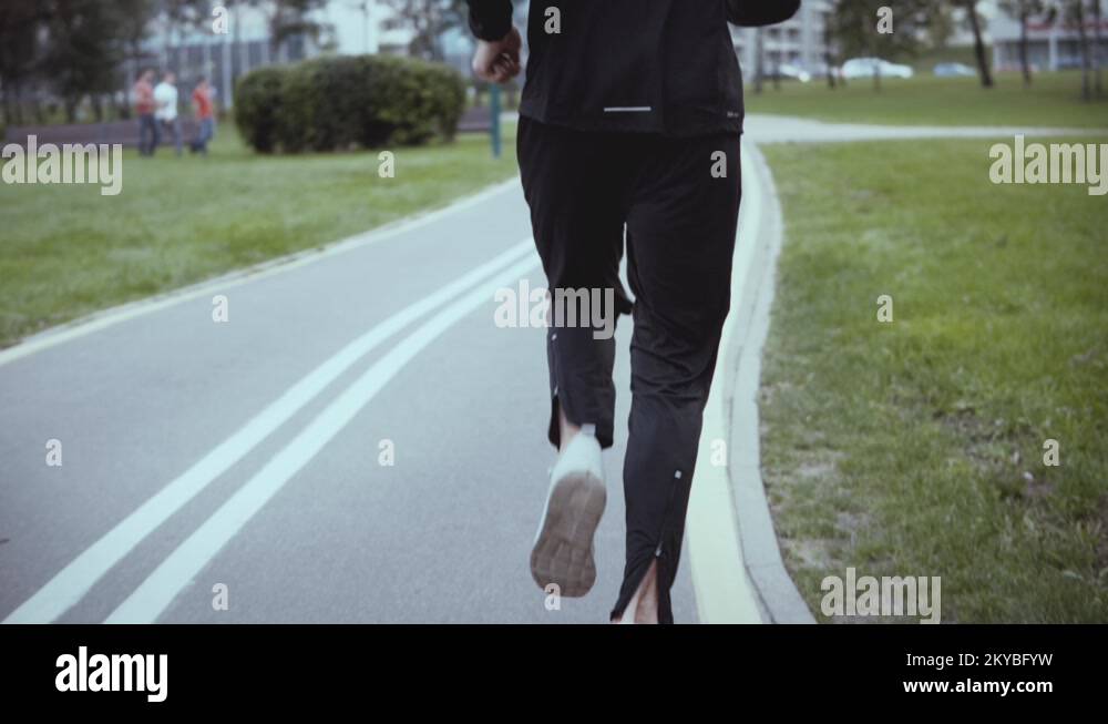 Man running along a city park road. Slow motion. Back view. Tilt up ...