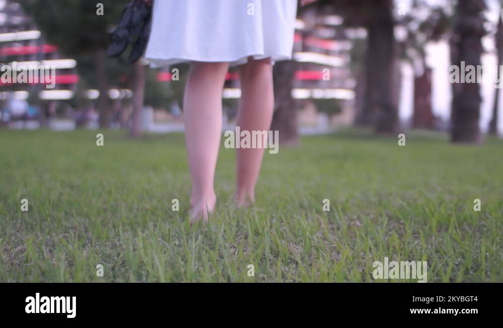 women legs go barefoot on the green grass. girl in white dress carries ...