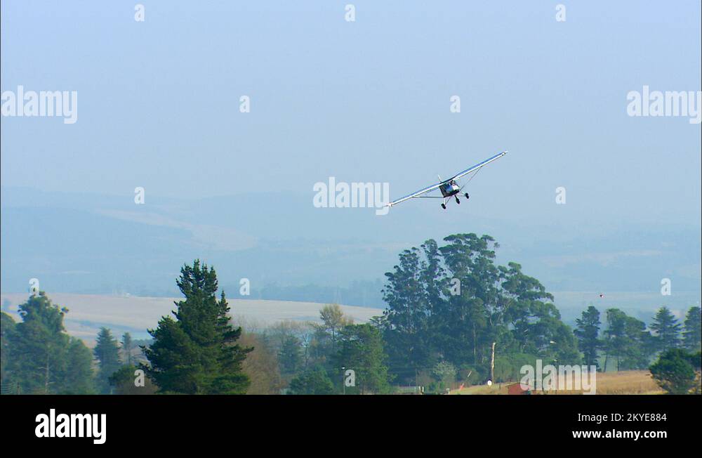 Light aircraft flying low over field with trees and small buildings ...