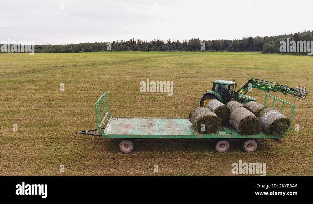 Large yellow field with green trailer loaded by hay bales and working ...