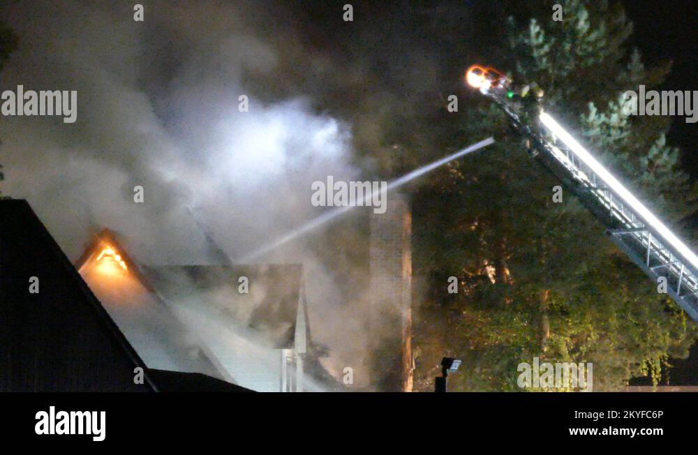 Fireman atop a fire ladder puts water on house with glow from flame ...