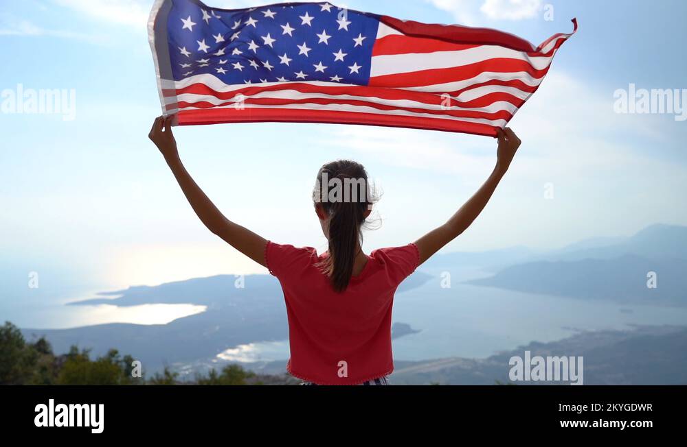 Child girl is waving American flag on top of mountain at sky background ...