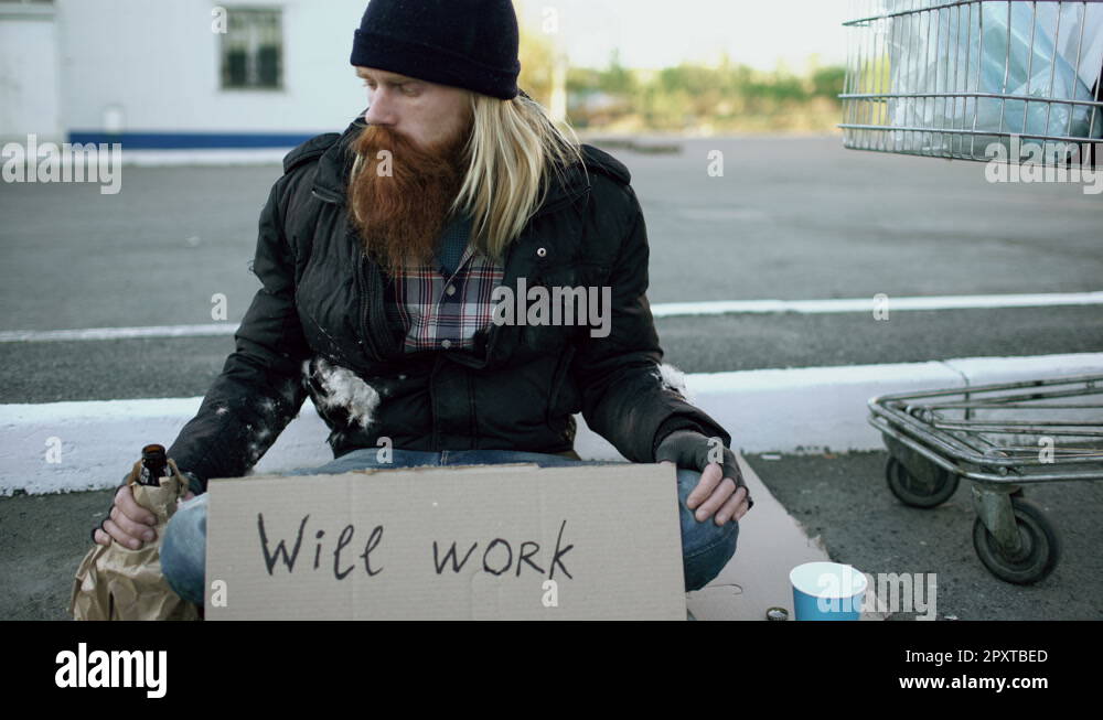 Tilt up of young homeless man with cardboard sitting near shopping cart ...