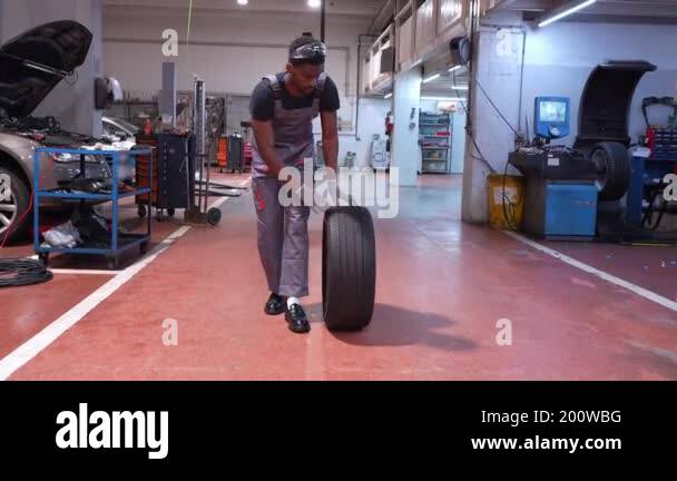 Skilled mechanic rolling heavy tire across workshop floor during ...