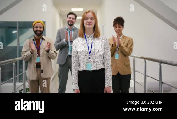 Red-haired young businesswoman crosses arms while employees stand behind in office corridor ...