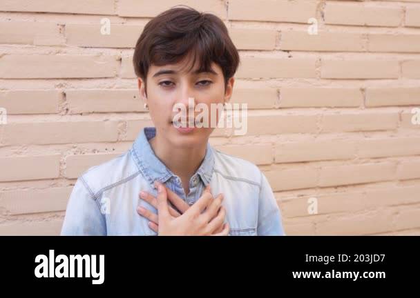 A young individual stands in front of a muted brick wall, expressing a ...