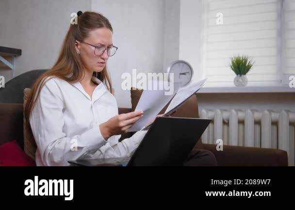 Young businesswoman in glasses sitting on sofa, comparing documents ...