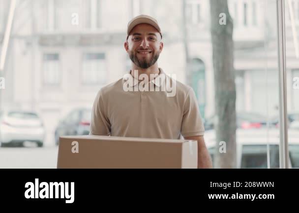 Friendly courier man with a joyful expression delivering a large cardboard box, wearing a beige ...