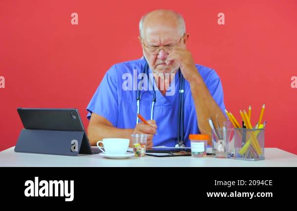 An elderly 60-year-old doctor, on a red background, works in an office ...