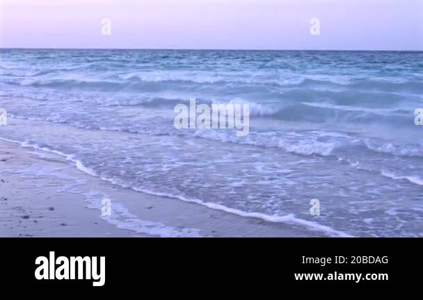 Gentle ocean waves capped in white wash over sandy beach under pastel ...